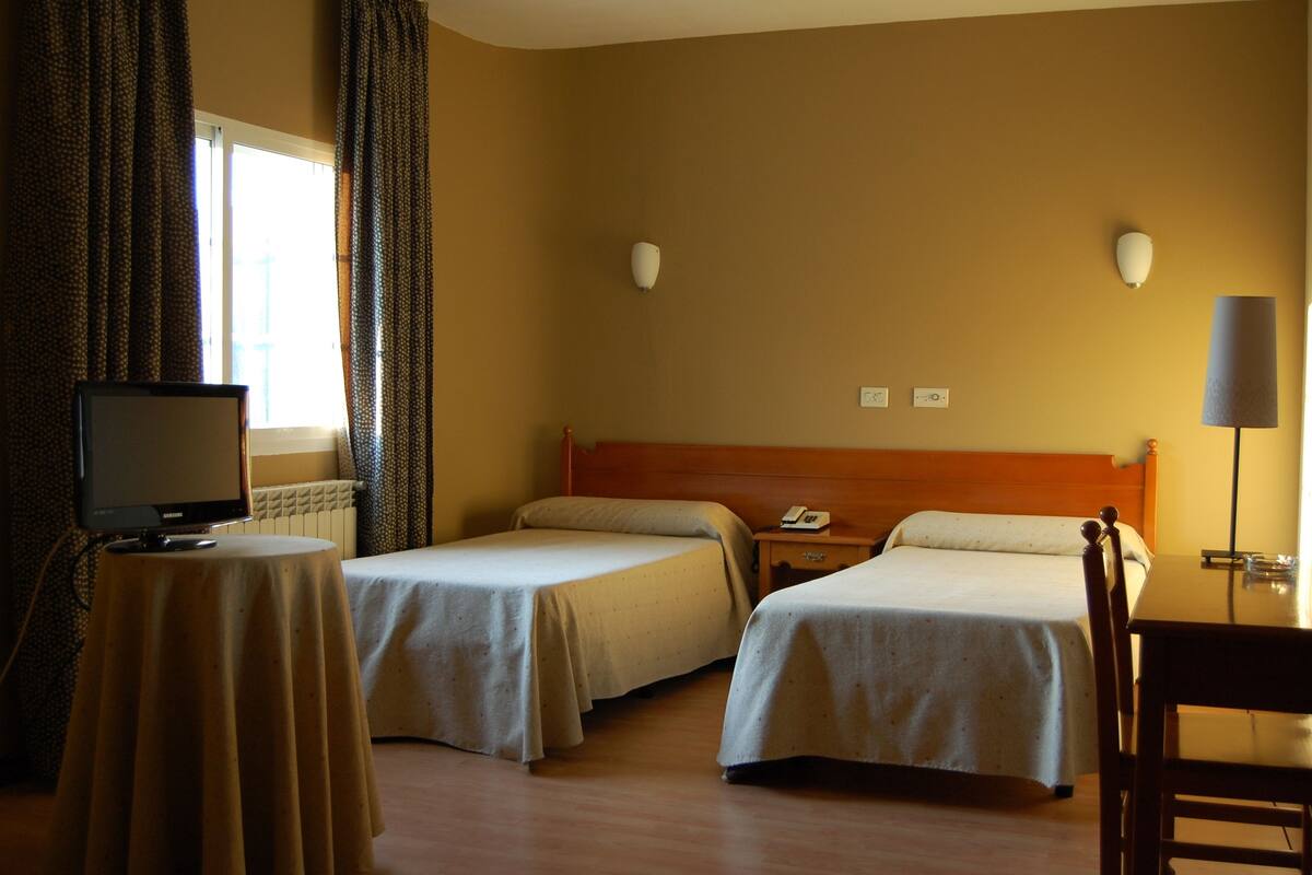 Quarto de hotel com duas camas de solteiro, uma mesa pequena com televisão, mesa de cabeceira com abajur e uma janela com cortinas.