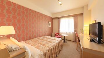 Quarto de hotel com cama, mesa de cabeceira com luminária e telefone, televisão, pequena mesa com cadeira e janela com cortinas.