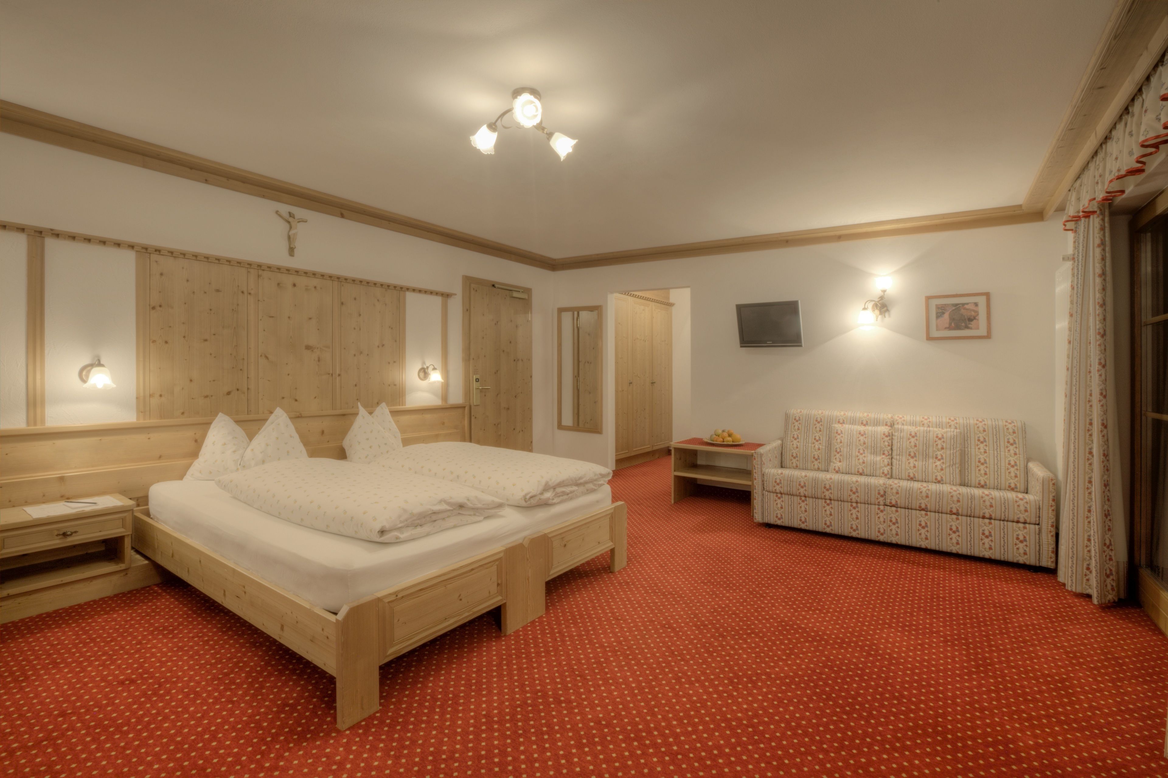 Quarto com cama de madeira, um sofá, mesa de cabeceira e televisão fixada na parede.