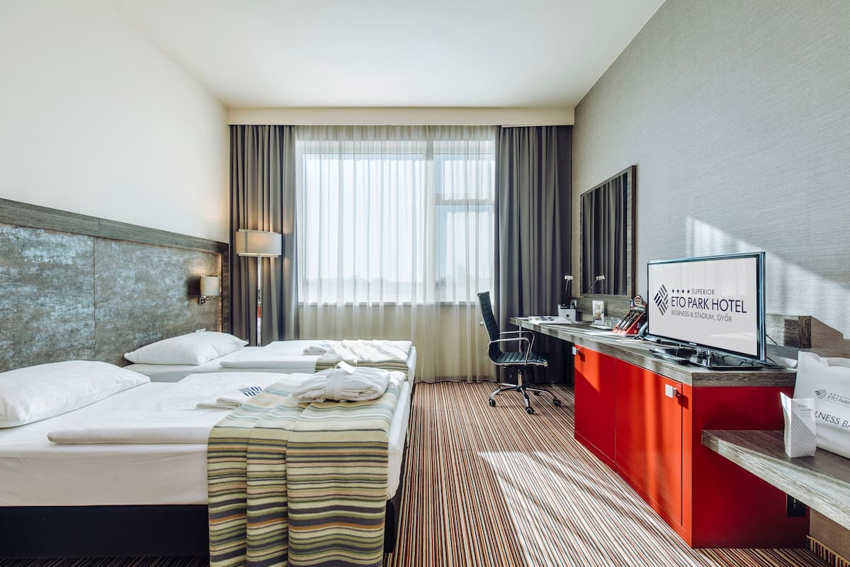 Quarto de hotel com duas camas, uma mesa de trabalho, televisão e vista da cidade através de amplas janelas.
