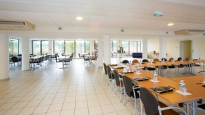 Uma sala de conferências ampla, com várias mesas e cadeiras, janelas grandes e teto com iluminação embutida.
