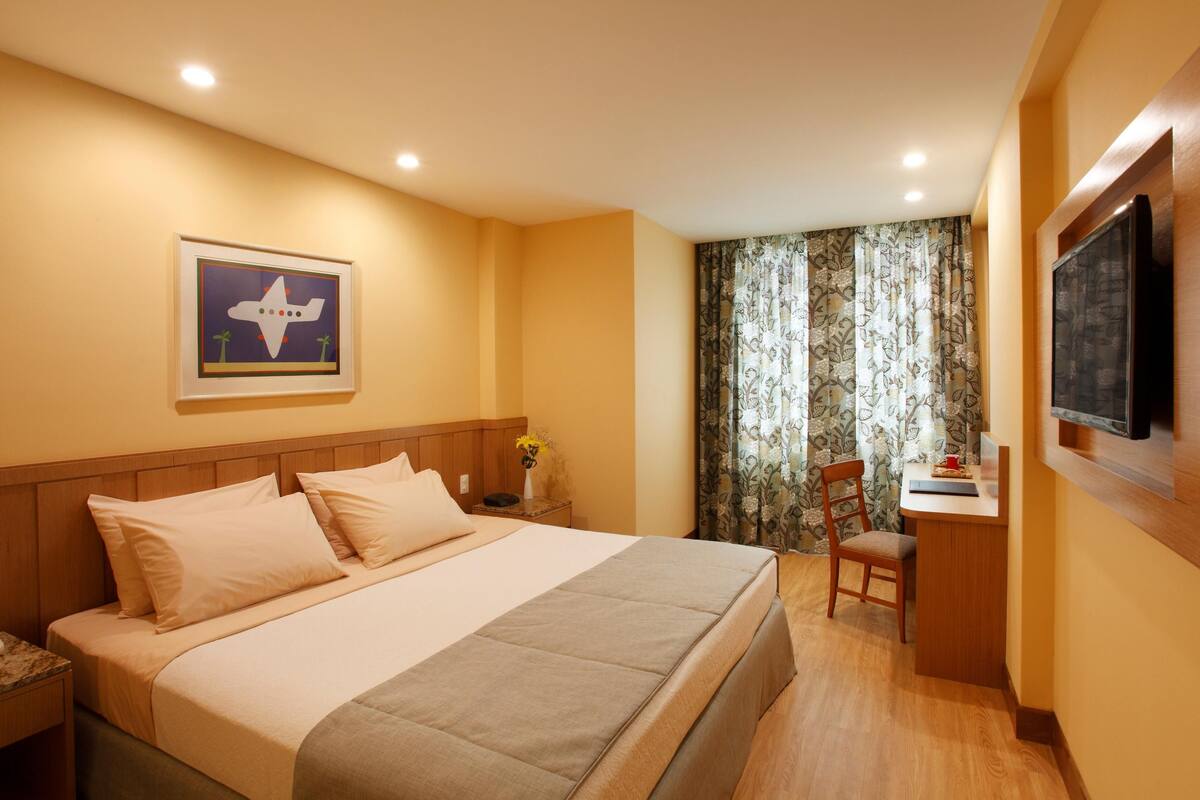 Quarto de hotel com cama, escrivaninha, cadeira, televisão e um quadro na parede.