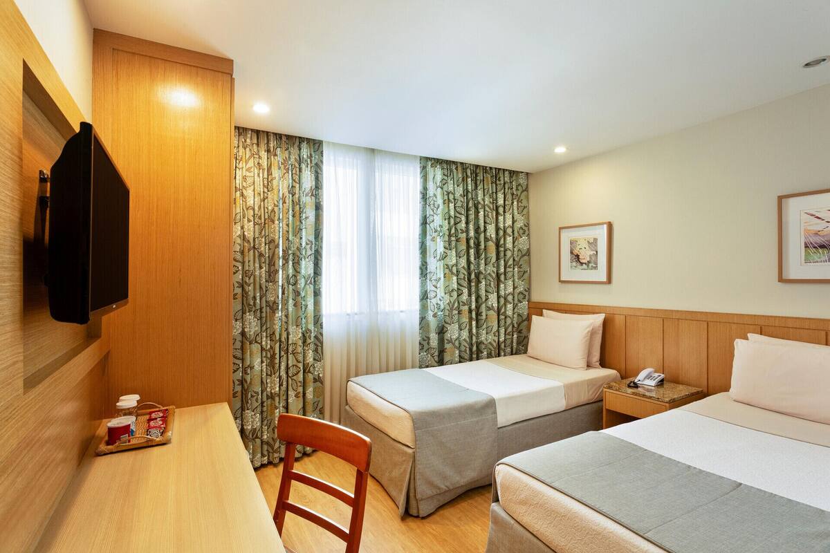 Quarto de hotel com duas camas, uma escrivaninha, uma cadeira, uma televisão e um telefone.
