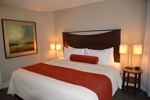 Quarto de hotel com uma cama grande, duas luminárias de cabeceira, um quadro na parede e uma mesinha.