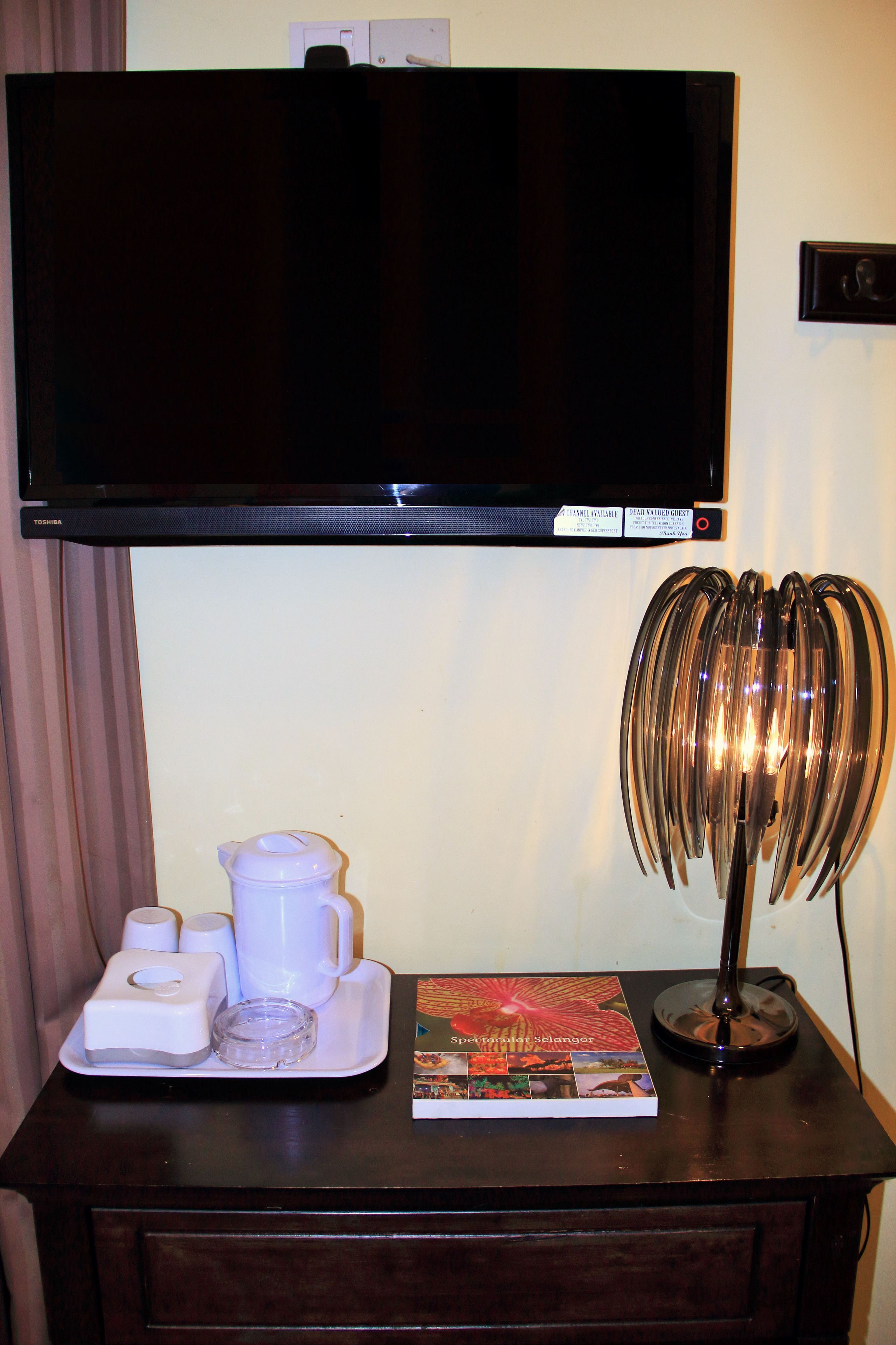 Uma mesa de cabeceira escura de madeira com uma televisão de tela plana, um abajur decorativo, uma bandeja com cafeteira, xícaras e um livro.