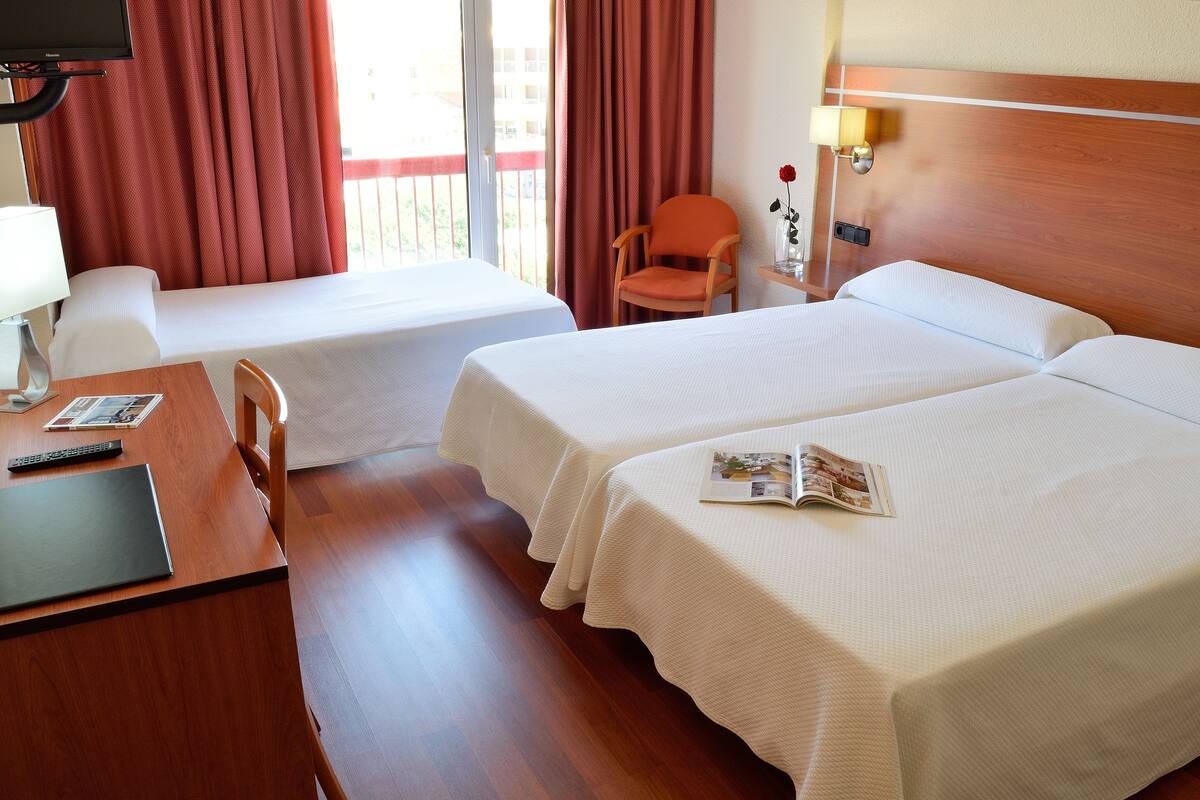 Quarto de hotel com duas camas, uma escrivaninha de madeira, uma cadeira, uma televisão e uma janela com cortinas.