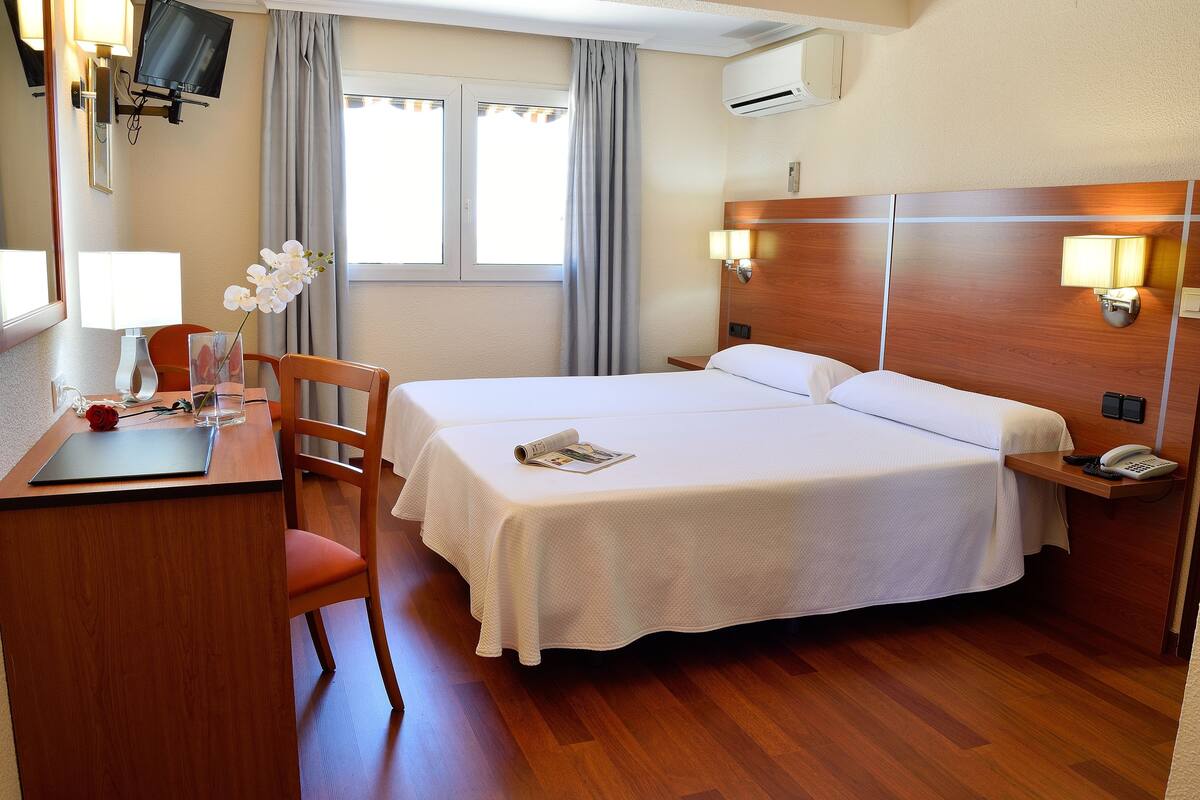 Quarto de hotel com cama, escrivaninha, cadeira e televisão fixada na parede.