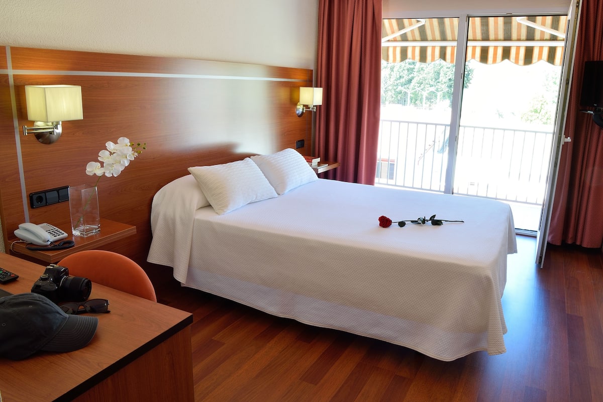 Quarto de hotel com cama, escrivaninha, telefone, câmera e vista para o exterior.