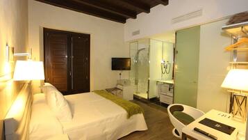 Quarto de hotel com cama, escrivaninha, cadeira, televisão, área de chuveiro e guarda-roupa.