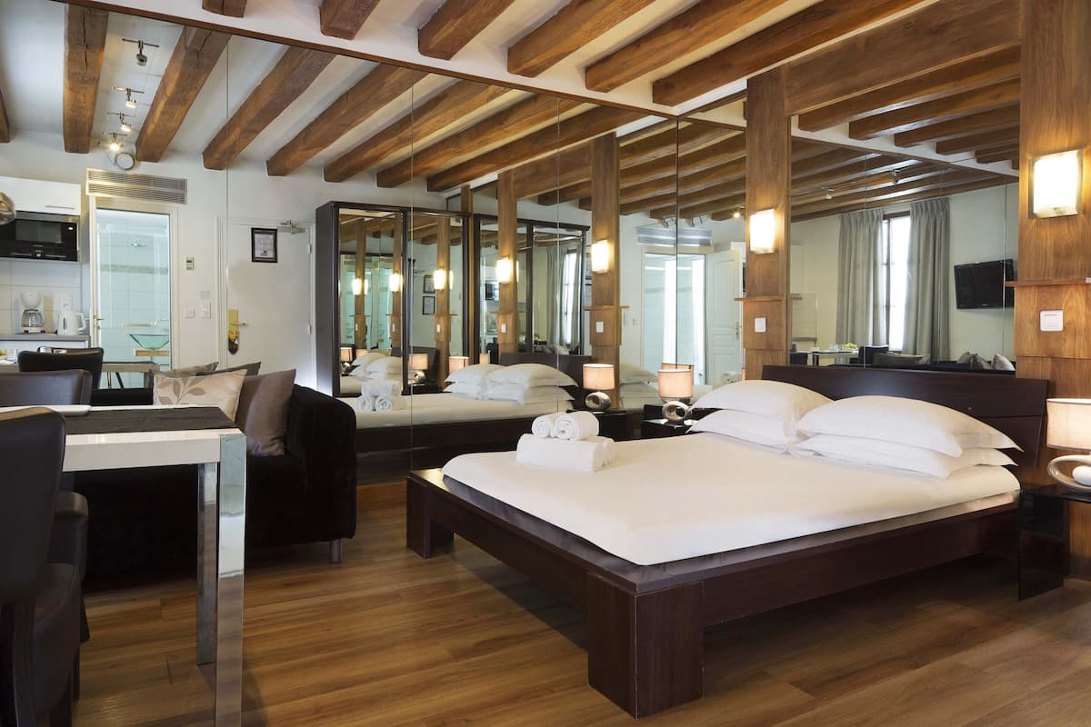 Quarto de hotel com teto de madeira, uma cama grande com roupa de cama branca, uma escrivaninha com cadeira e um espelho refletindo o quarto.