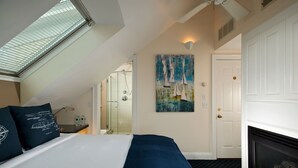 Quarto de hotel com uma cama grande, um claraboia, um quadro de barcos a vela, uma lareira e um espaço com chuveiro.