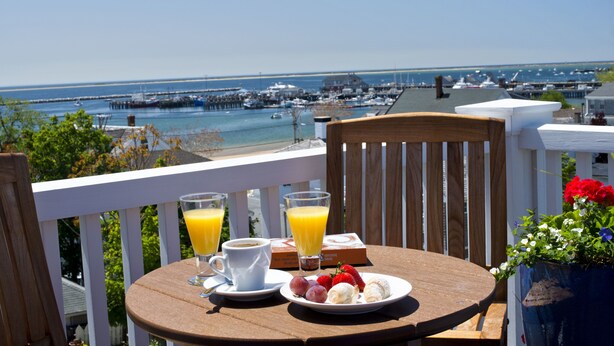 Uma mesa de madeira com itens de café da manhã e bebidas em uma varanda com vista para um porto com barcos e um cais.