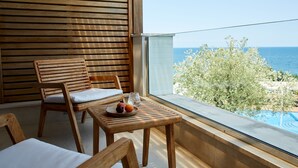 Varanda com mobiliário de madeira, uma mesa com frutas e um copo, e vista para o mar e vegetação.
