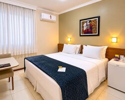 Quarto de hotel com cama, mesas de cabeceira, escrivaninha e janela com cortinas.