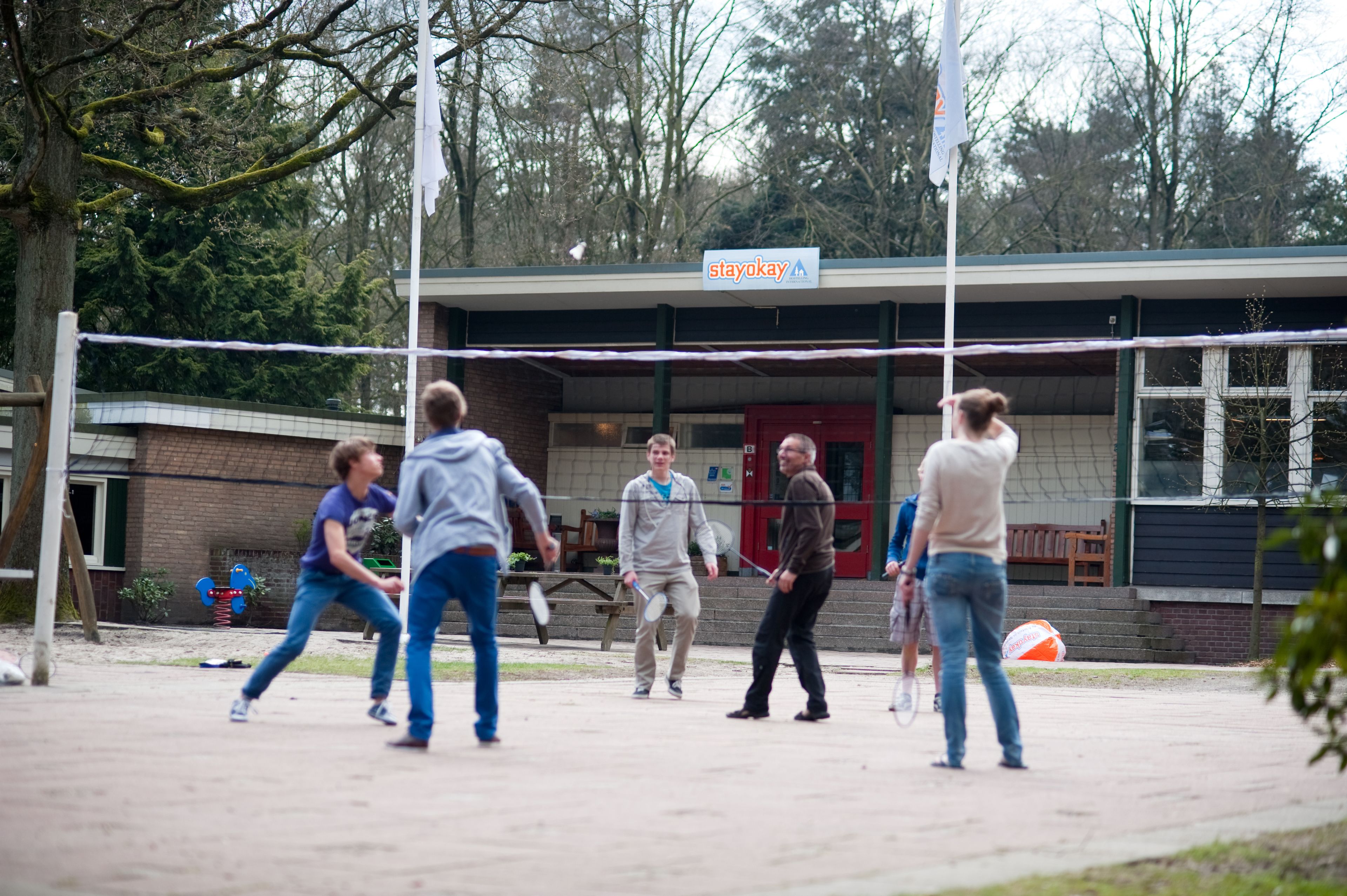 Um grupo de pessoas jogando badminton em área externa, com um edifício ao fundo.