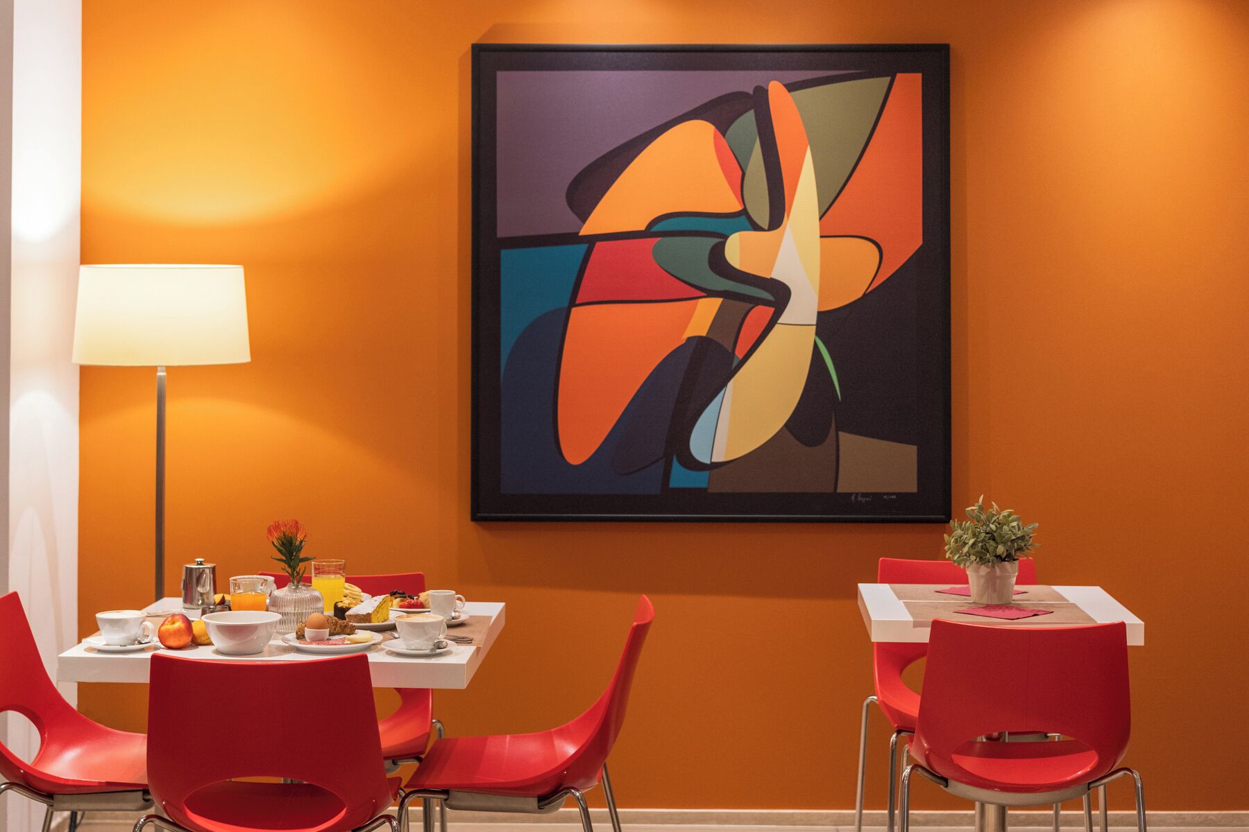 Uma área de refeições com mesa posta para café da manhã, cadeiras vermelhas e um quadro abstrato moderno na parede.