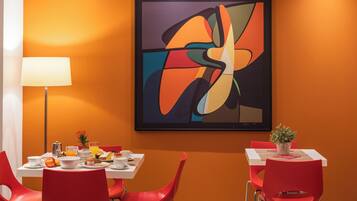 Uma área de refeições com mesa posta para café da manhã, cadeiras vermelhas e um quadro abstrato moderno na parede.