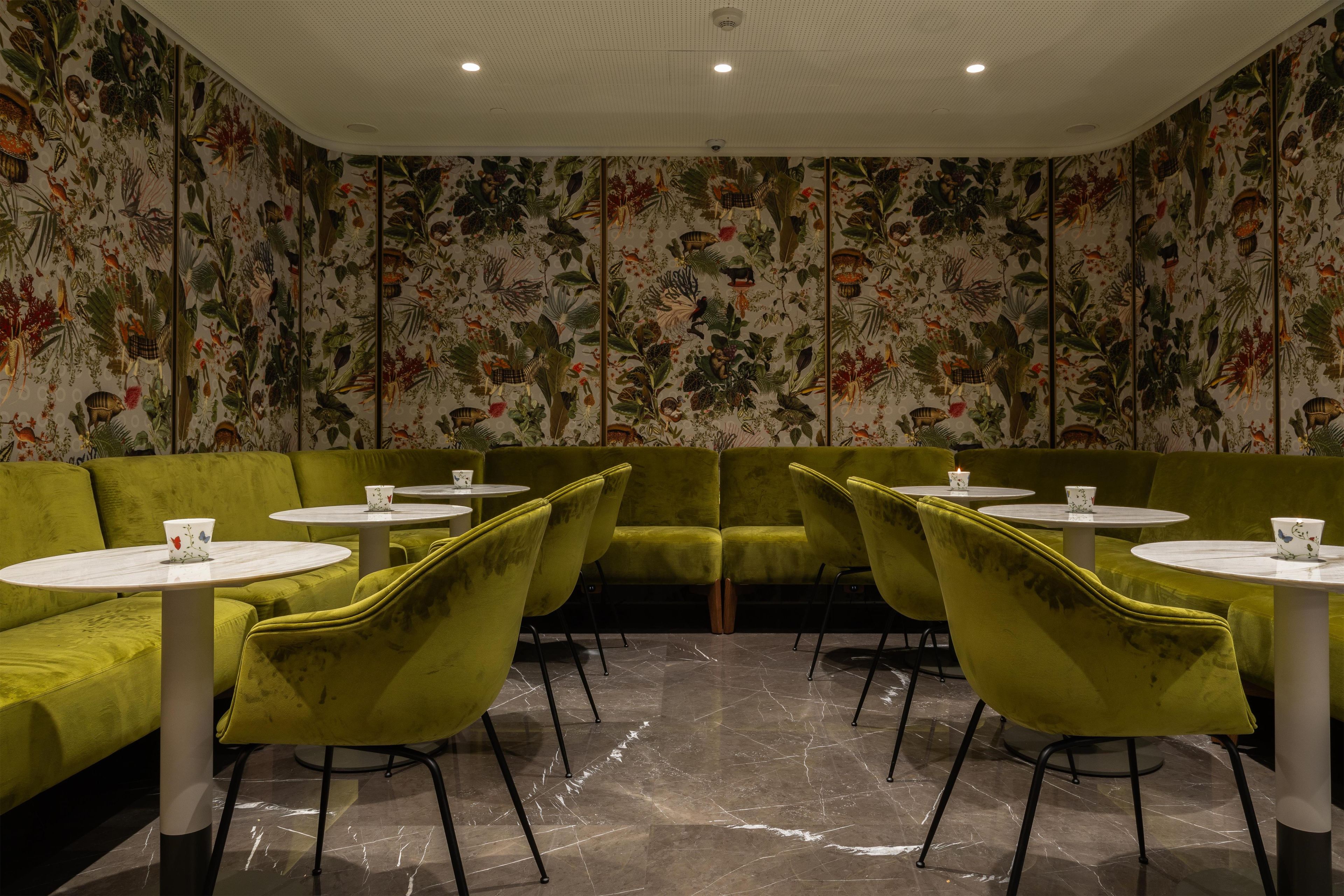 Interior de restaurante com assentos de veludo verde, mesas redondas brancas e um papel de parede com padrão floral.