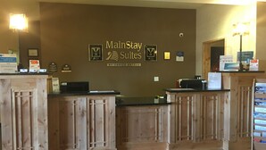 Uma área de recepção com painéis de madeira, um letreiro da MainStay Suites by Choice Hotels e diversos avisos e comodidades.