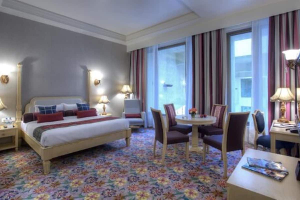 Quarto de hotel com cama, escrivaninha, duas cadeiras, uma mesinha e janela com cortinas.