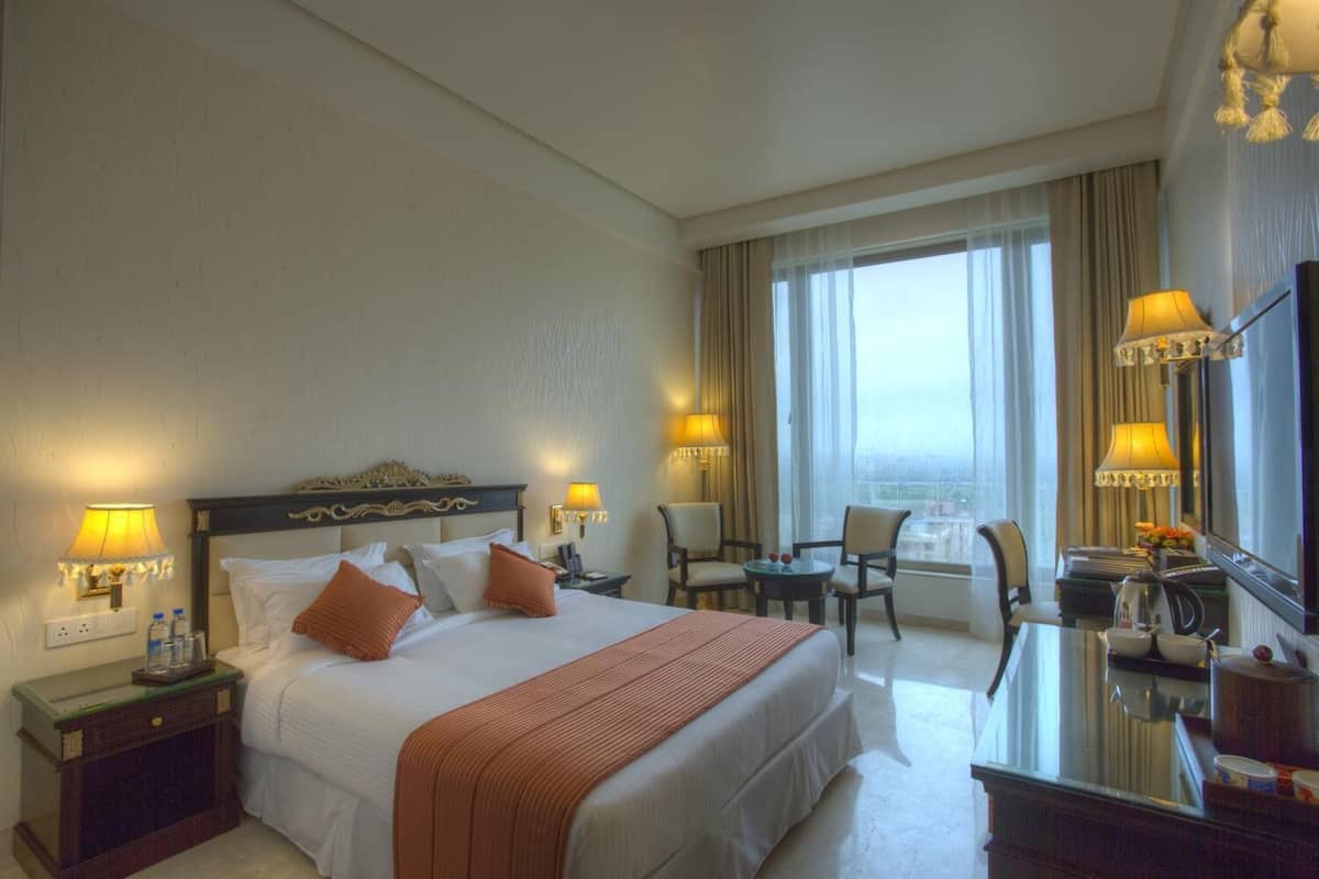 Quarto de hotel com uma cama grande, duas mesas de cabeceira, uma escrivaninha com cadeira, uma janela ampla com cortinas e um espelho.