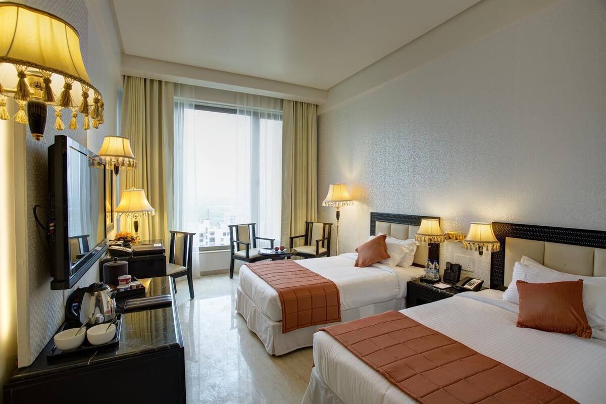 Quarto de hotel com duas camas, uma escrivaninha com cadeira, televisão e uma janela grande com cortinas.