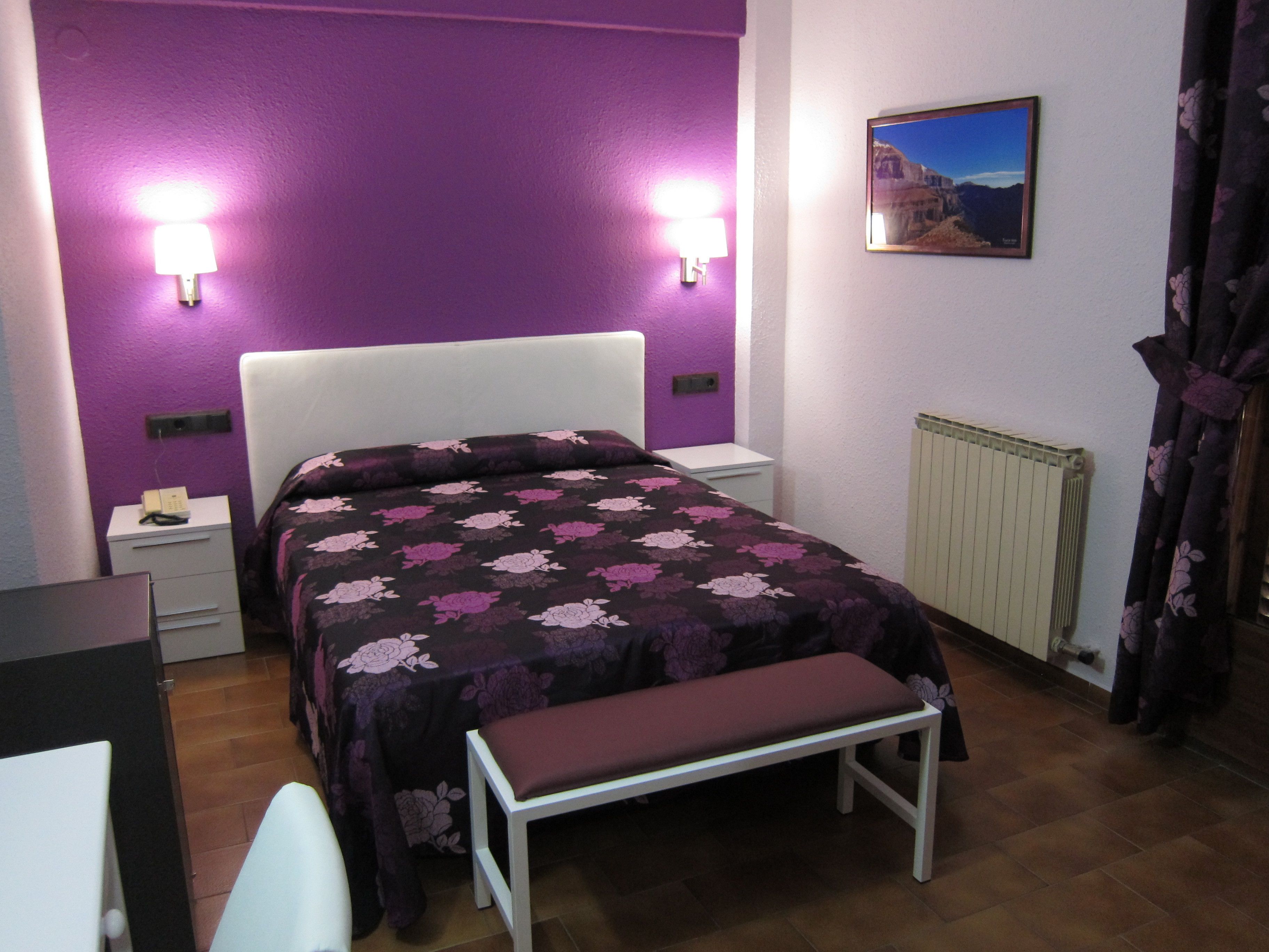 Quarto com cama, mesas de cabeceira, escrivaninha, um banquinho e um radiador.
