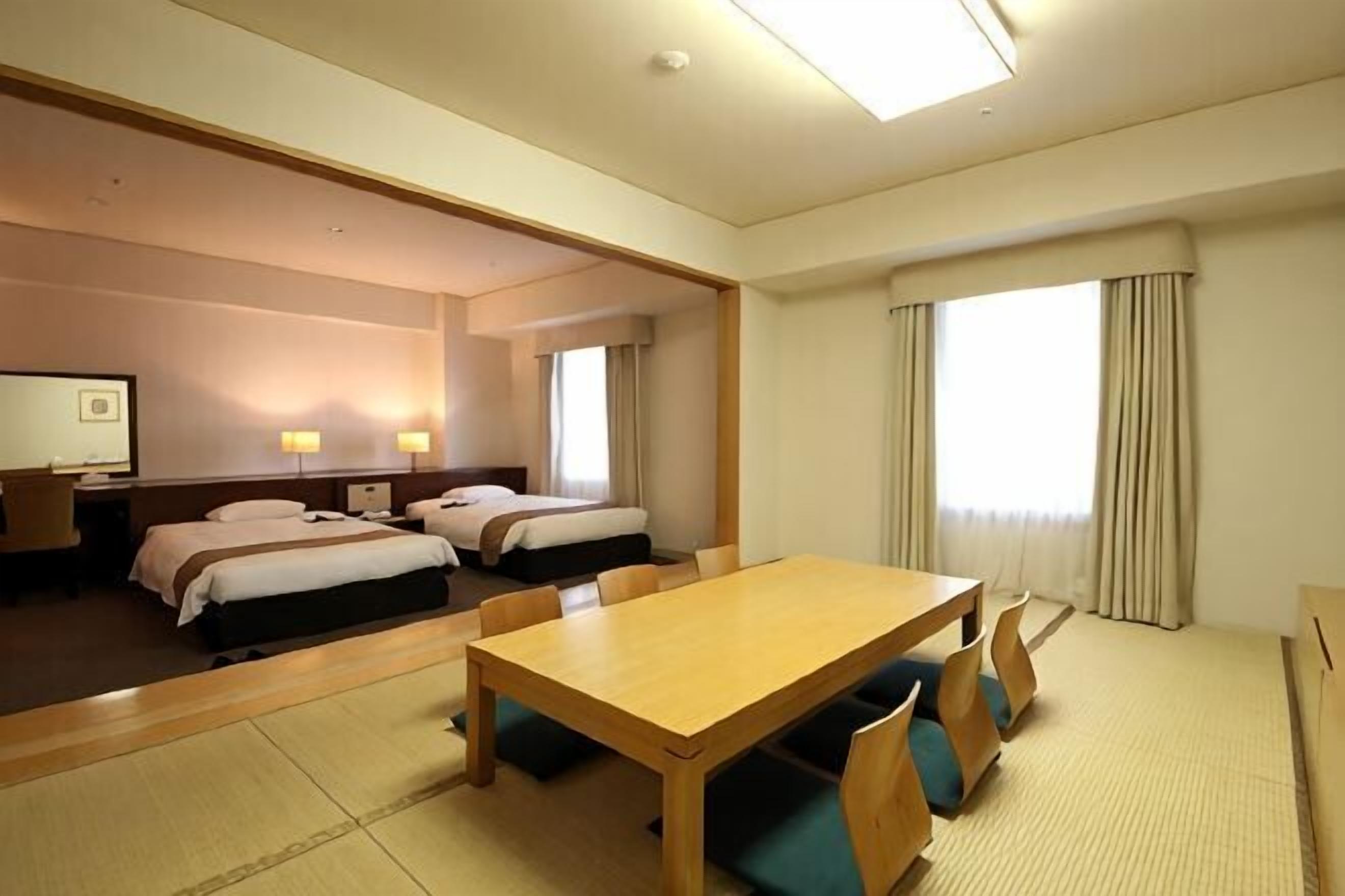 Quarto de hotel com mesa de jantar de madeira, duas camas, uma televisão e uma janela grande.
