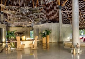 Um hall espaçoso com teto alto, pilares de concreto e telhado de palha. Há cadeiras de madeira e plantas em vasos.