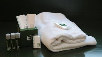 Uma toalha branca dobrada, um conjunto de produtos de higiene, incluindo um sabonete em barra e três frascos, e uma caixa verde com um logotipo sobre uma superfície escura.