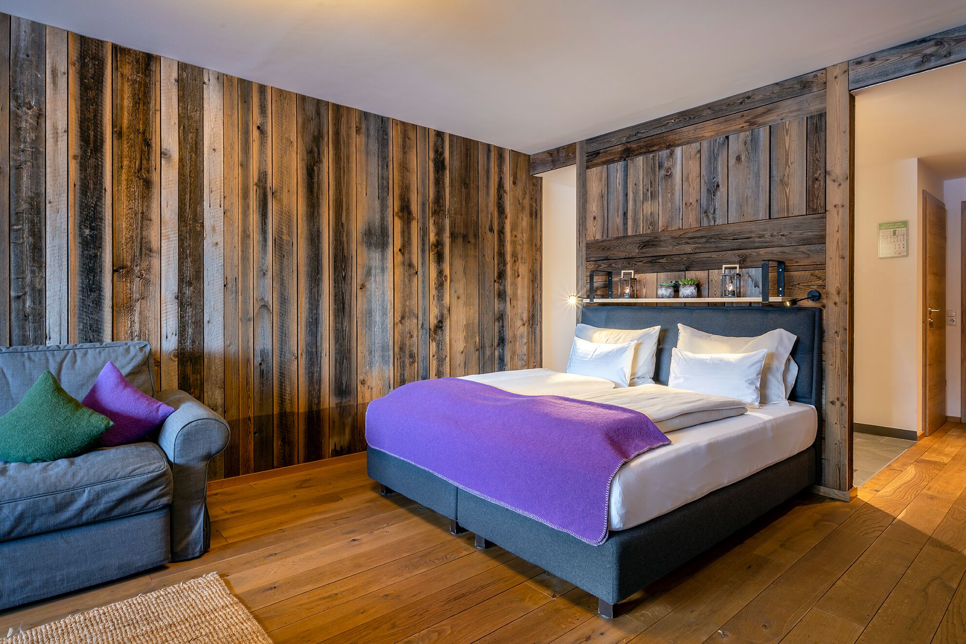 Quarto com parede de madeira, cama com roupa de cama branca e roxa, sofá cinza com almofadas coloridas e piso de madeira.