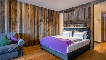 Quarto com parede de madeira, cama com roupa de cama branca e roxa, sofá cinza com almofadas coloridas e piso de madeira.