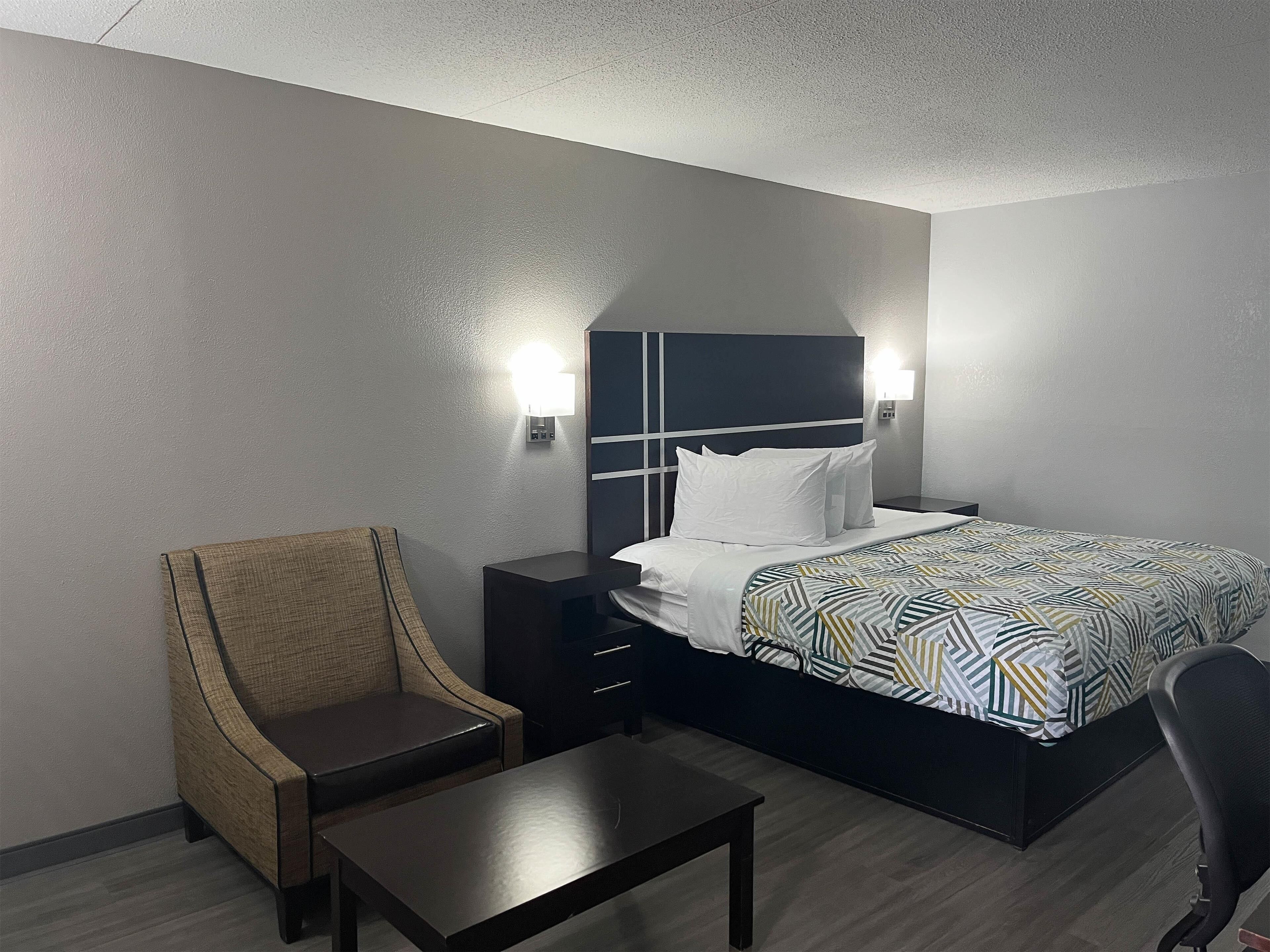 Quarto de hotel com cama, uma cadeira, uma mesinha e um criado-mudo.