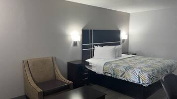 Quarto de hotel com cama, uma cadeira, uma mesinha e um criado-mudo.
