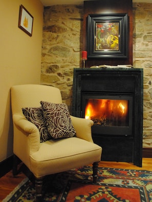 Um quarto aconchegante com parede de pedra, lareira com um quadro emoldurado acima, poltrona com estampa de bolinhas e almofada padronizada, e um tapete colorido no chão.