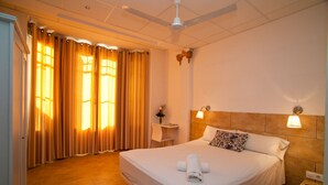 Quarto de hotel com uma cama grande, uma mesa de cabeceira, um ventilador de teto e uma janela com cortinas.