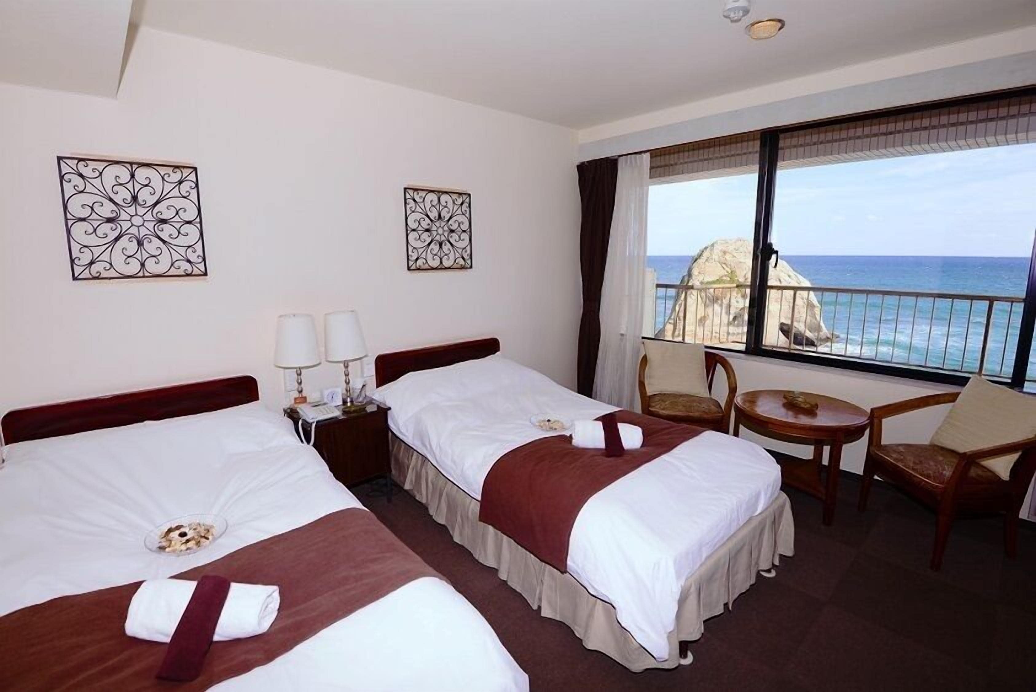 Quarto de hotel com duas camas, uma mesinha, uma cadeira e vista para o mar.