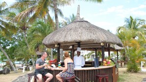 Bar ao ar livre com telhado de palha, um cozinheiro, um homem e uma mulher sentados.