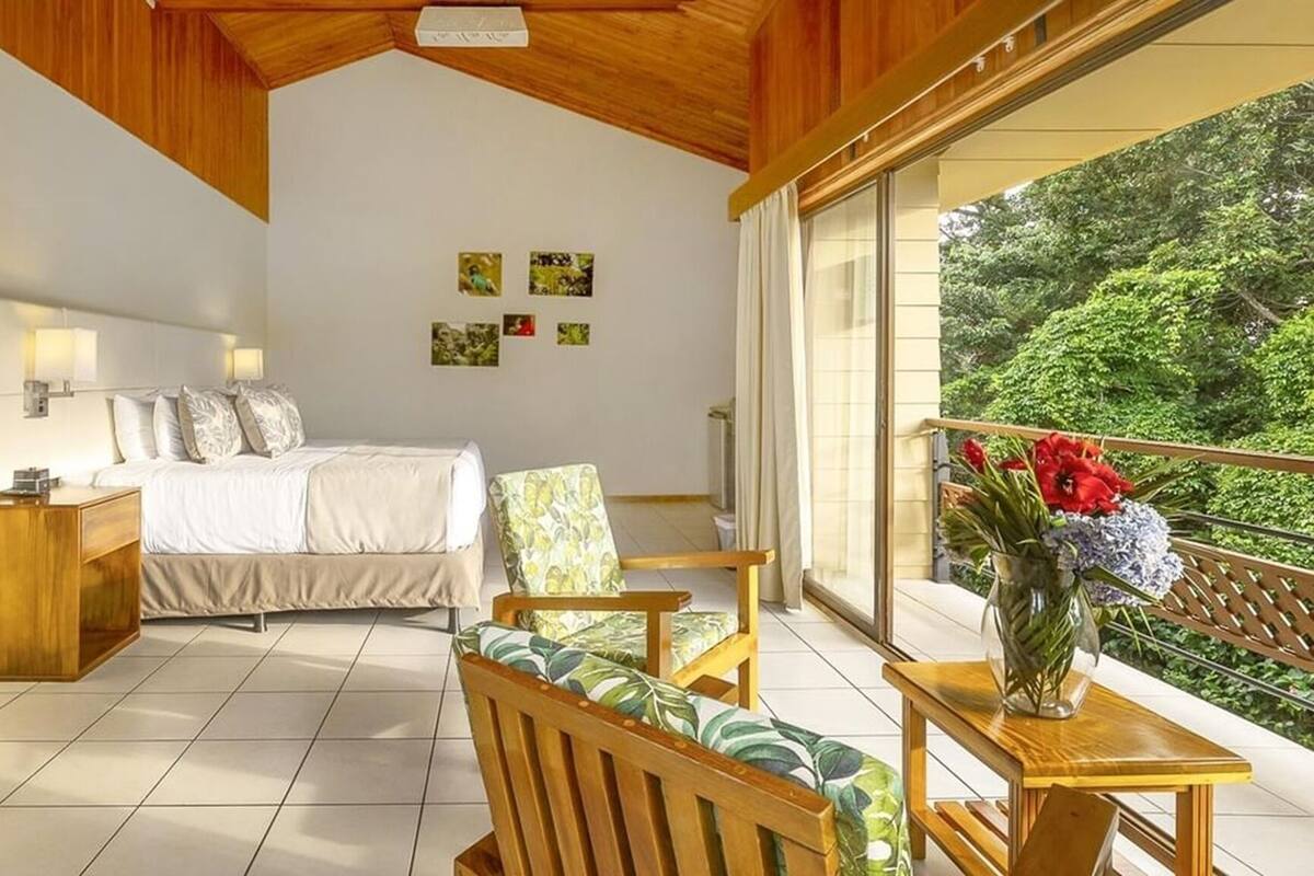 Quarto com cama, mesa de cabeceira, cadeira e varanda com vista para o verde.