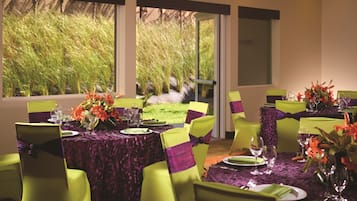 Uma sala de jantar com mesas preparadas para quatro pessoas, cadeiras com estofamento verde, toalhas de mesa roxas e centros de mesa com flores.