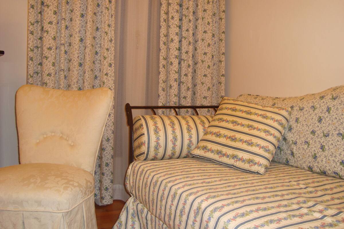 Quarto com cama de padrão floral, almofada combinando e uma cadeira.