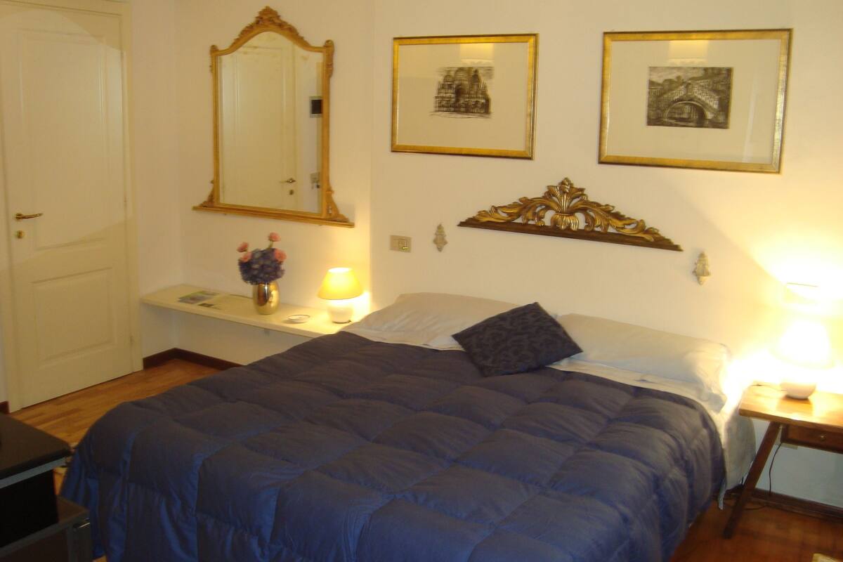 Quarto com cama, mesa de cabeceira, abajur, espelho e duas quadros pendurados na parede.