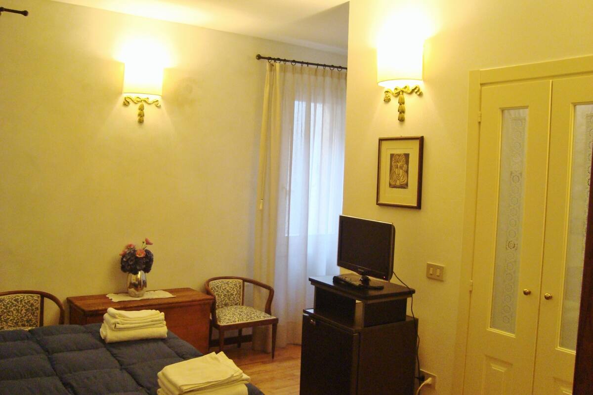 Quarto de hotel com uma cama, uma mesa com um computador, uma cadeira, uma televisão e uma porta.