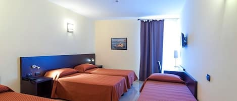 Quarto de hotel com quatro camas dispostas em linha, um ventilador de teto, uma mesa de cabeceira com abajur e um quadro na parede.