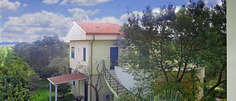 Uma casa de dois andares com telhado de telhas vermelhas, varanda coberta e jardim com diversas plantas.