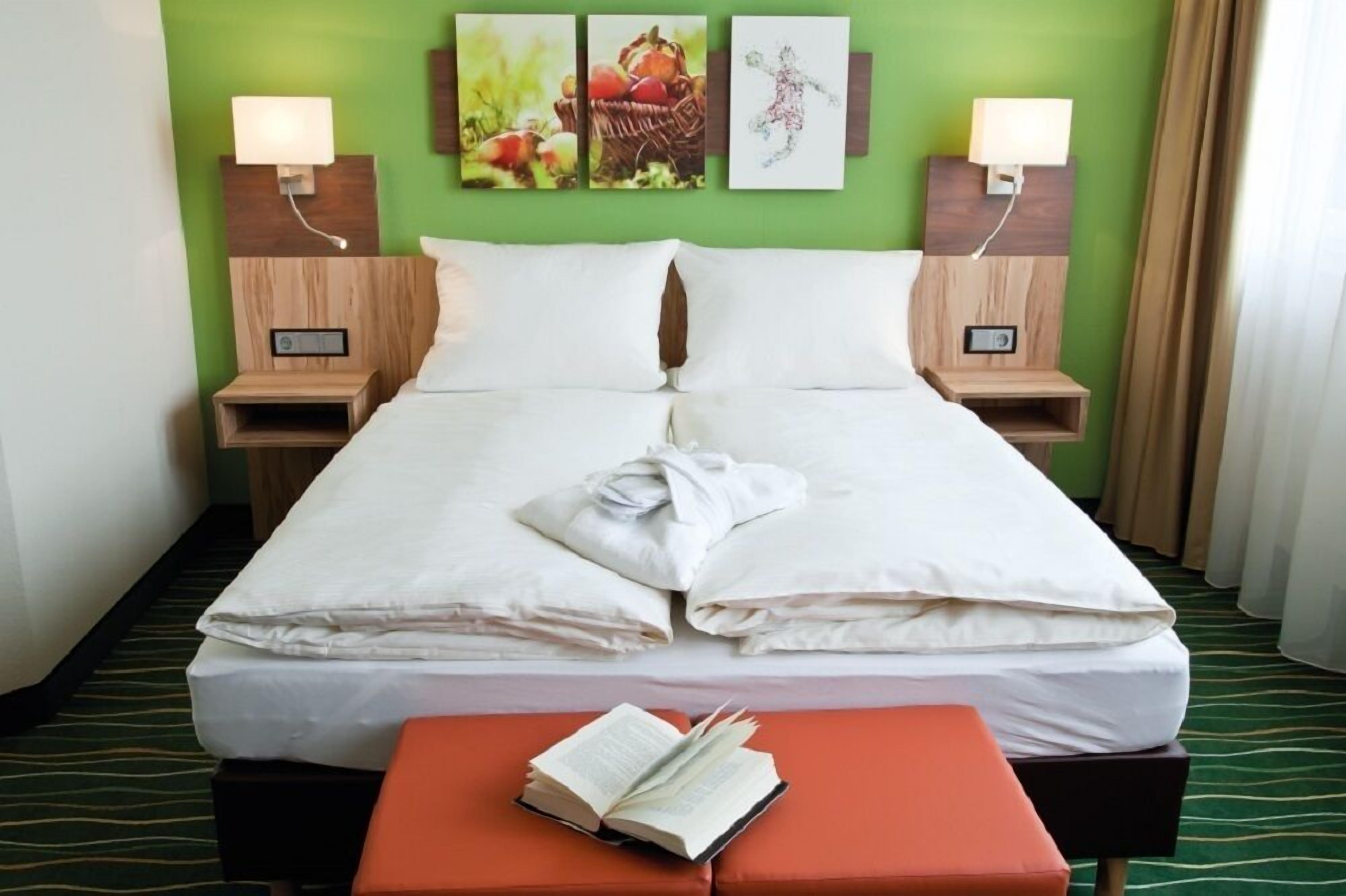 Quarto de hotel com uma cama bem arrumada, duas mesas de cabeceira com luminárias, uma parede verde com obras de arte e uma janela com cortinas.
