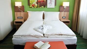 Quarto de hotel com uma cama bem arrumada, duas mesas de cabeceira com luminárias, uma parede verde com obras de arte e uma janela com cortinas.
