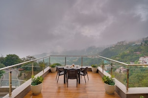 Um terraço com mesa e cadeiras, plantas em vasos e vista para uma paisagem montanhosa.