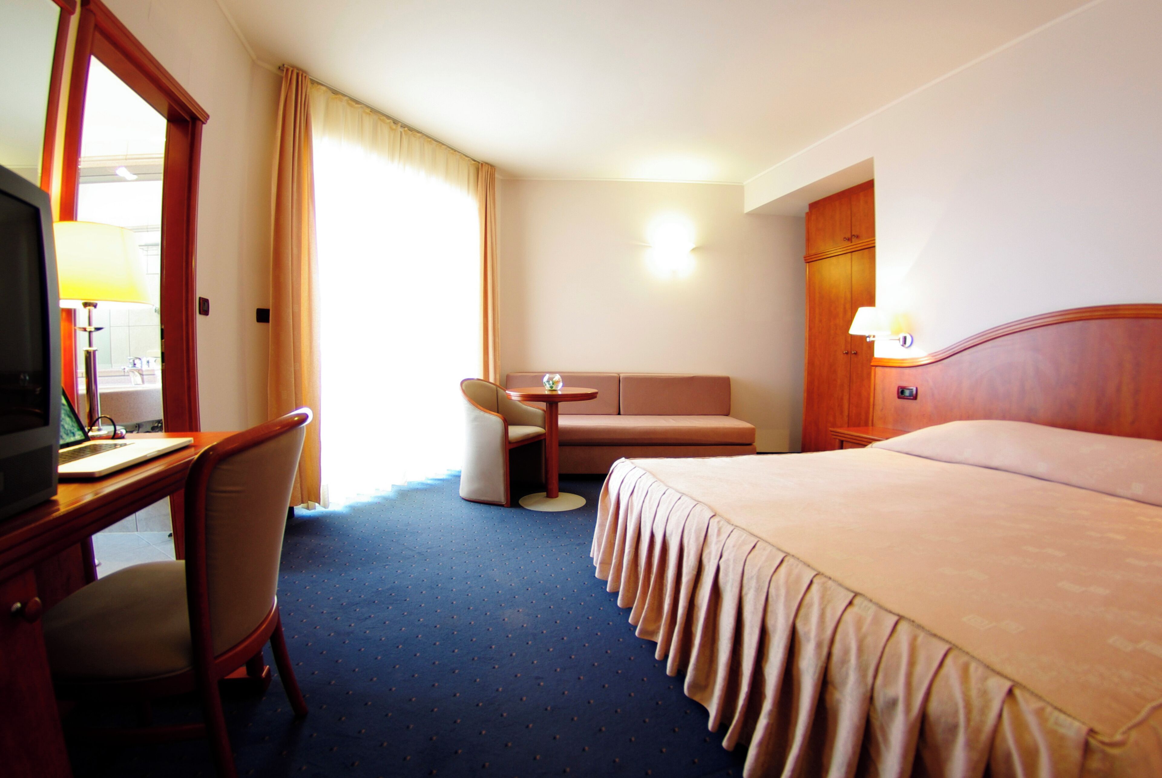 Quarto de hotel com uma cama, escrivaninha com computador, cadeira, mesa pequena e televisão.