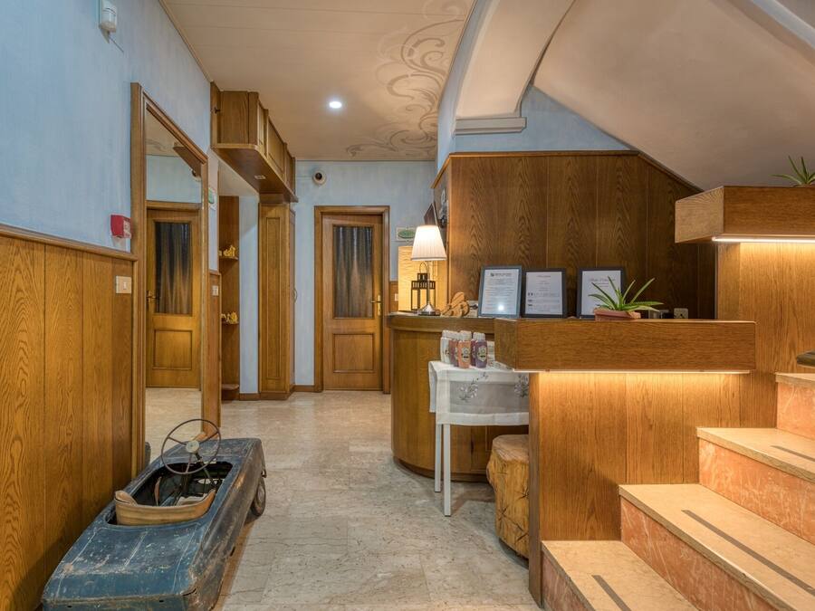 Um saguão de hotel com painéis de madeira, um balcão de recepção, uma pequena mesa com objetos e uma escadaria.
